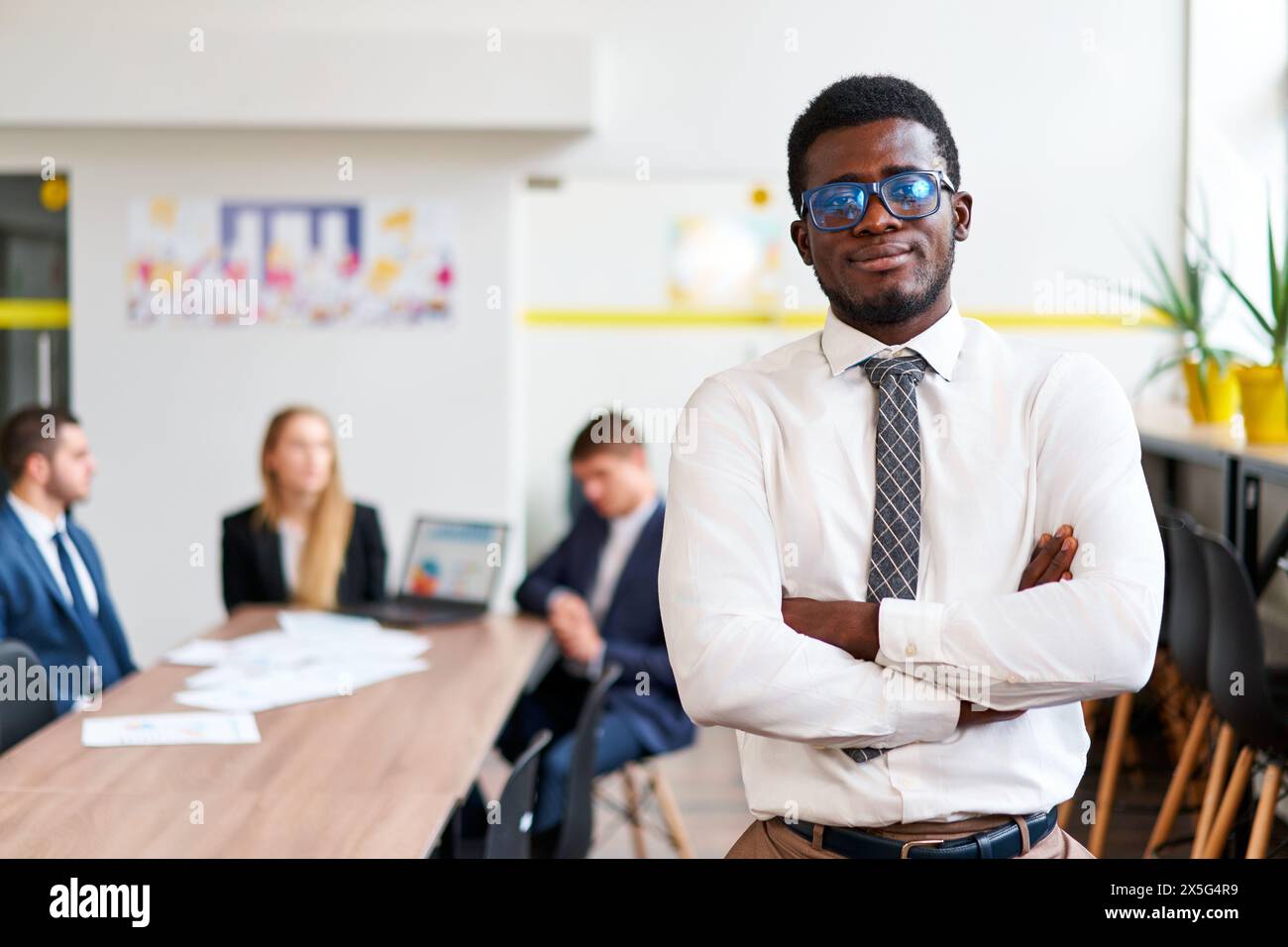 Confident African businessman stands in modern office, arms crossed. Teamwork in background ...