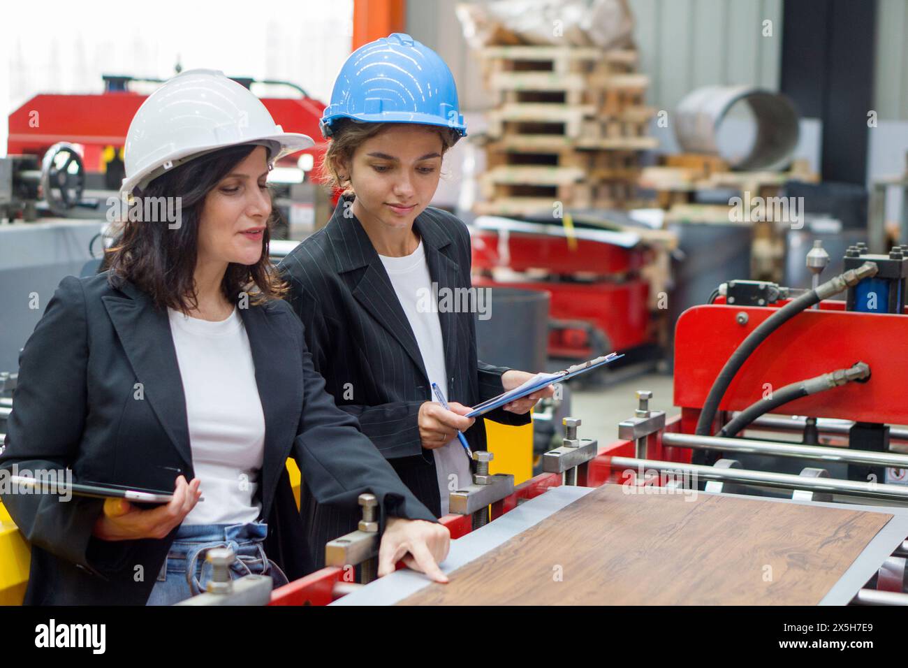 Two women dressed in black suit and safety helmet are inspecting work in a factory setting Stock ...