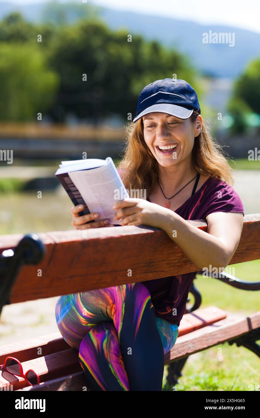 A joyful young woman with a book laughs while seated on a park bench ...
