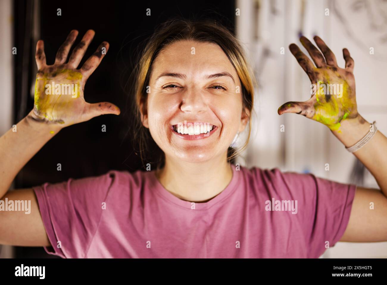 A happy female artist displays her colorful paint-stained hands with a broad smile in her ...