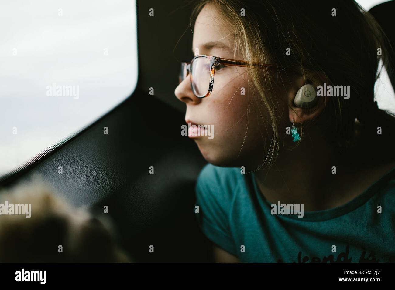 Tween girl in glasses looks out car window on road trip with headphone ...