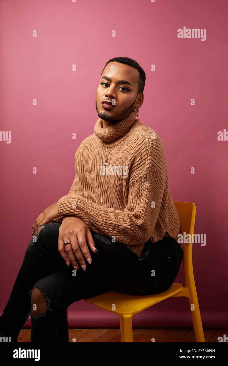 Studio portrait of queer man sitting on chair against purple background ...