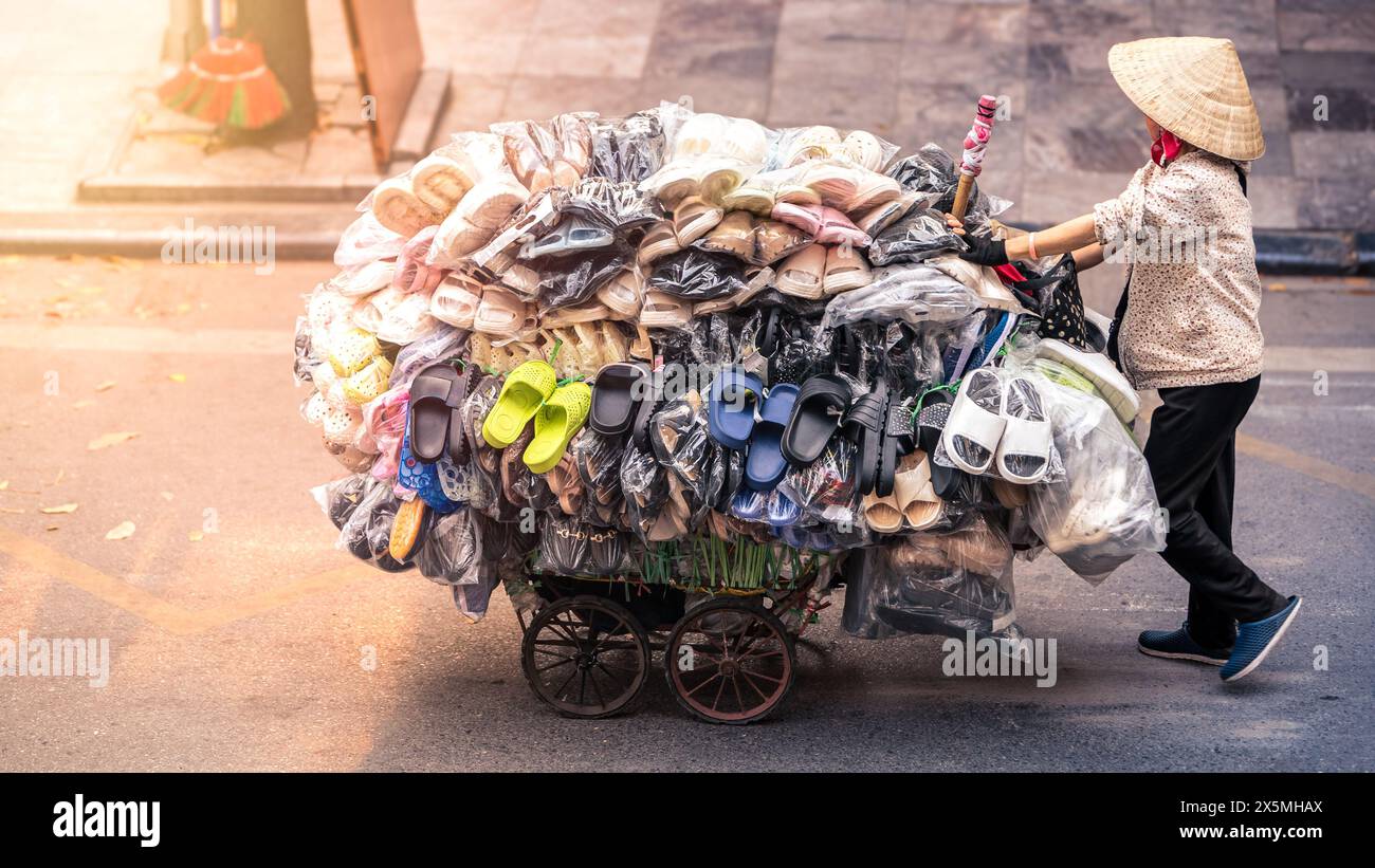 A street vendor wearing a straw hat, pushes a cart overflowing with ...