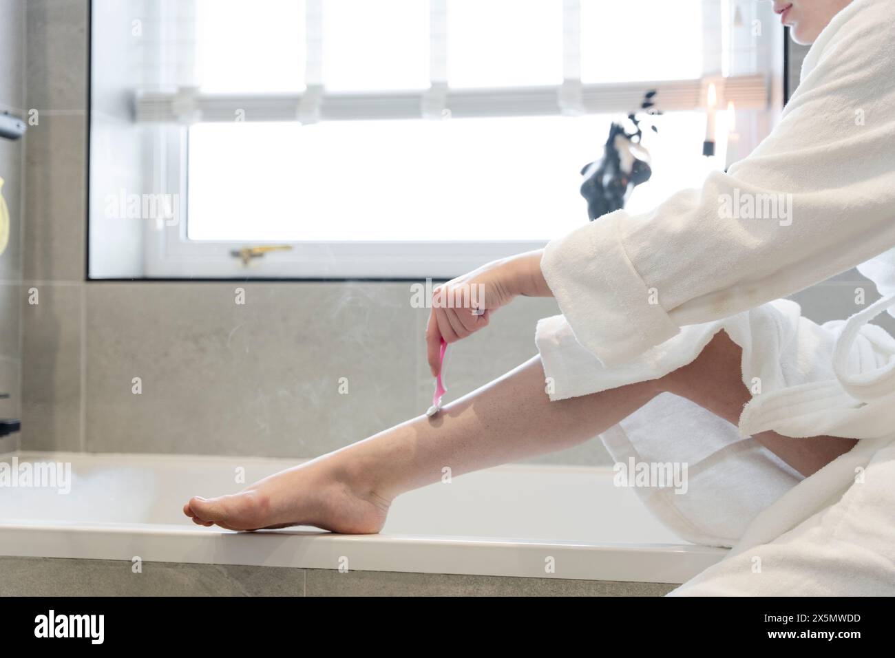 Woman in bathrobe shaving leg at bathtub edge Stock Photo - Alamy