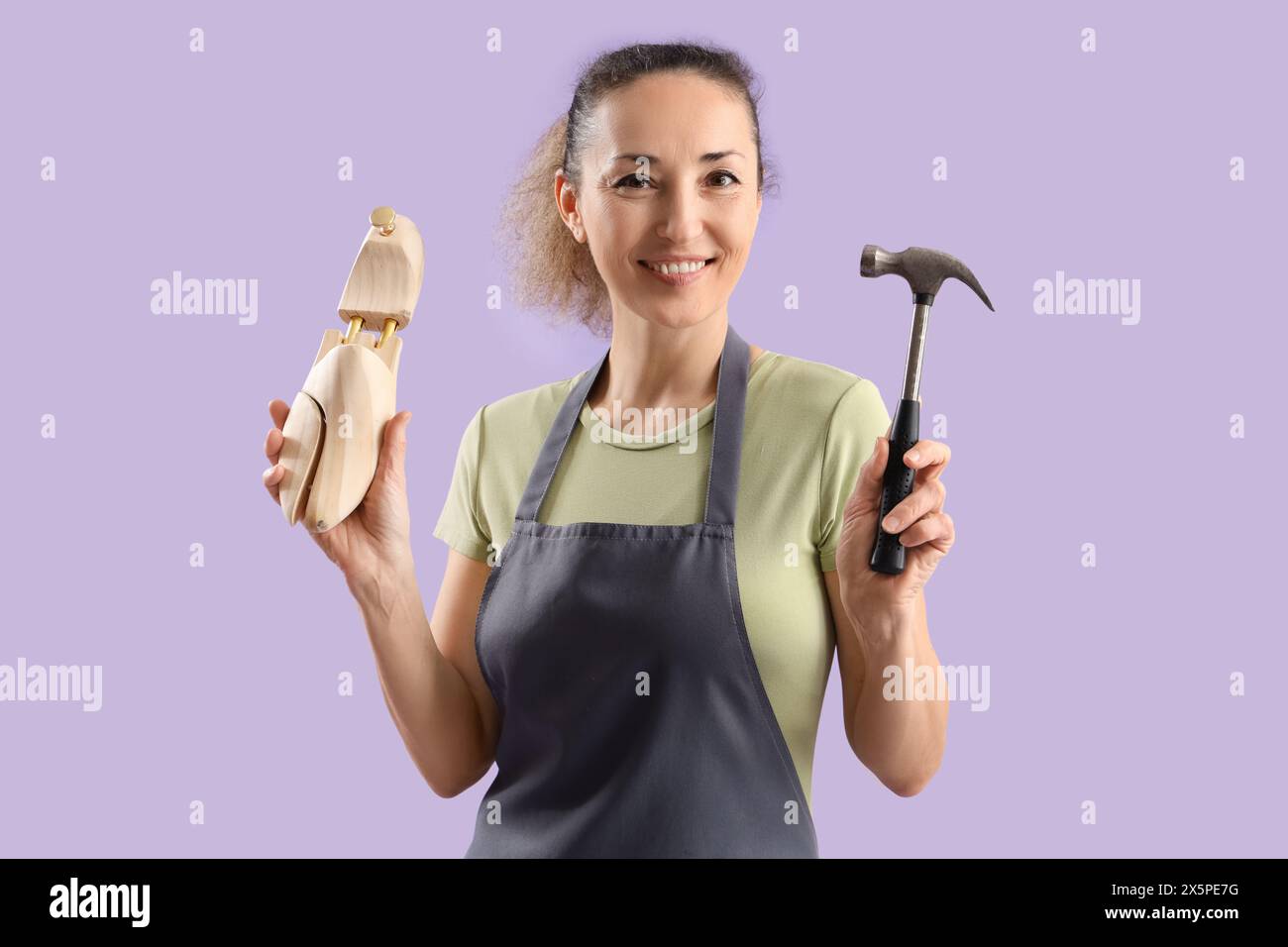 Happy female shoemaker with wooden shoe tree and hammer on lilac ...