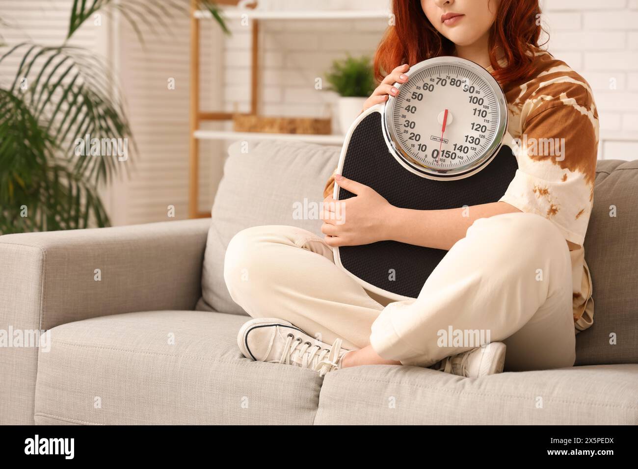 Young woman with weight scales sitting on sofa at home. Weight gain ...