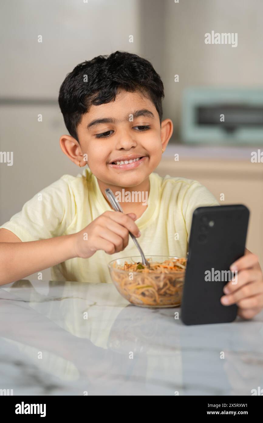 vertical shot of Indian preteen kid eating noodles while using mobile ...