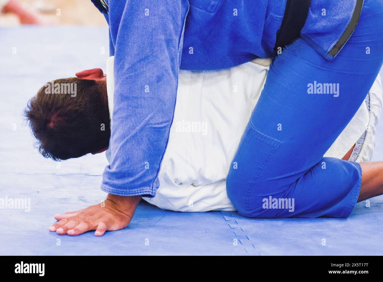Two men on a blue mat in a Brazilian Jiu-Jitsu training facility. He is ...