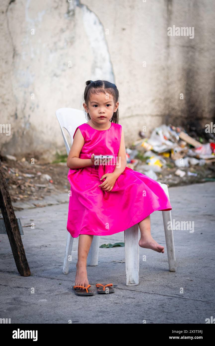 A young Filipino girl in a pretty pink dress looks at her phone outside ...