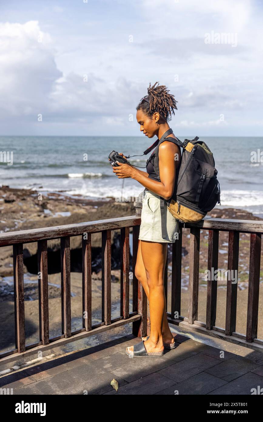 Indonesia, Bali, Female tourist standing with digital camera by sea ...