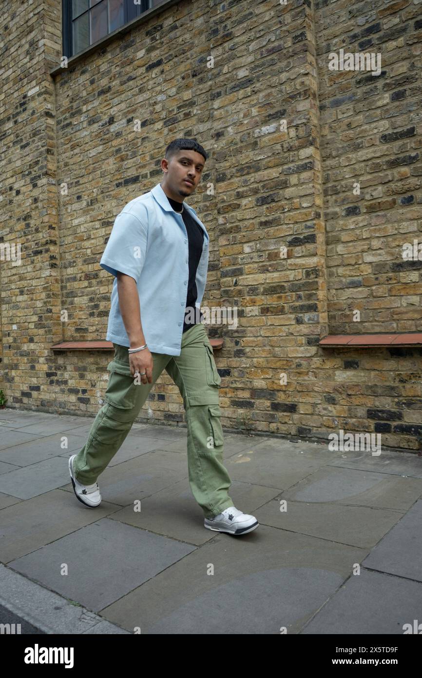 Man wearing unbuttoned shirt and cargo pants walking along brick wall ...