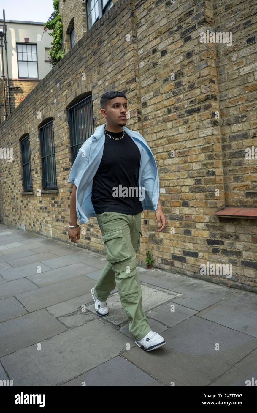 Man wearing unbuttoned shirt and cargo pants walking along brick wall ...