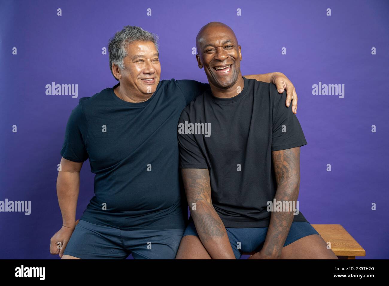 Portrait of two men laughing while sitting on bench against purple ...