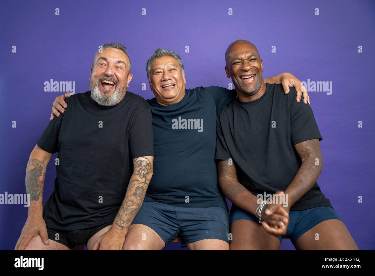 Portrait of three men laughing while sitting on bench against purple ...