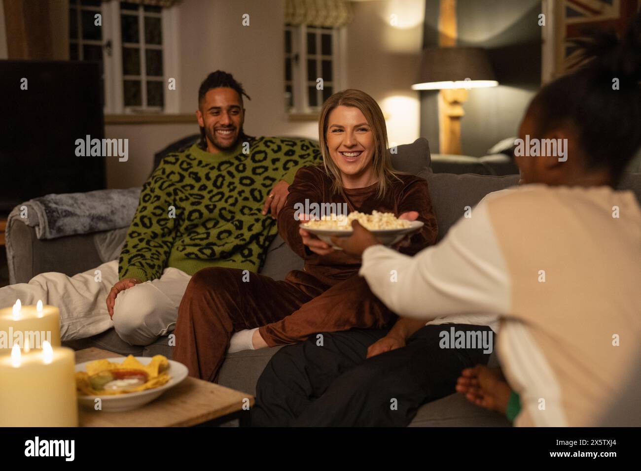 Smiling friends relaxing on sofa at home, sharing popcorn Stock Photo ...