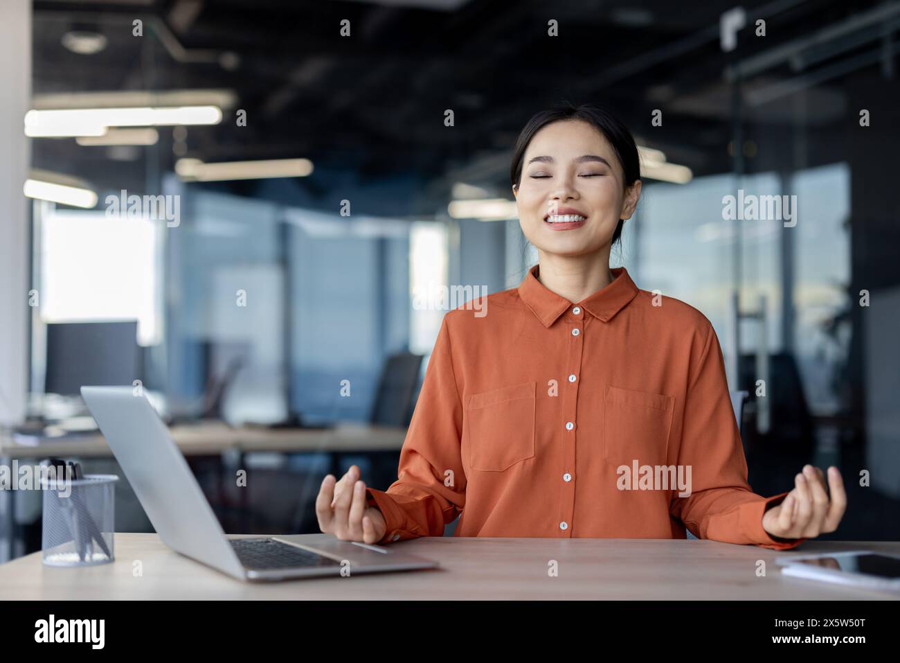 A professional Asian woman peacefully meditates in a bright office ...
