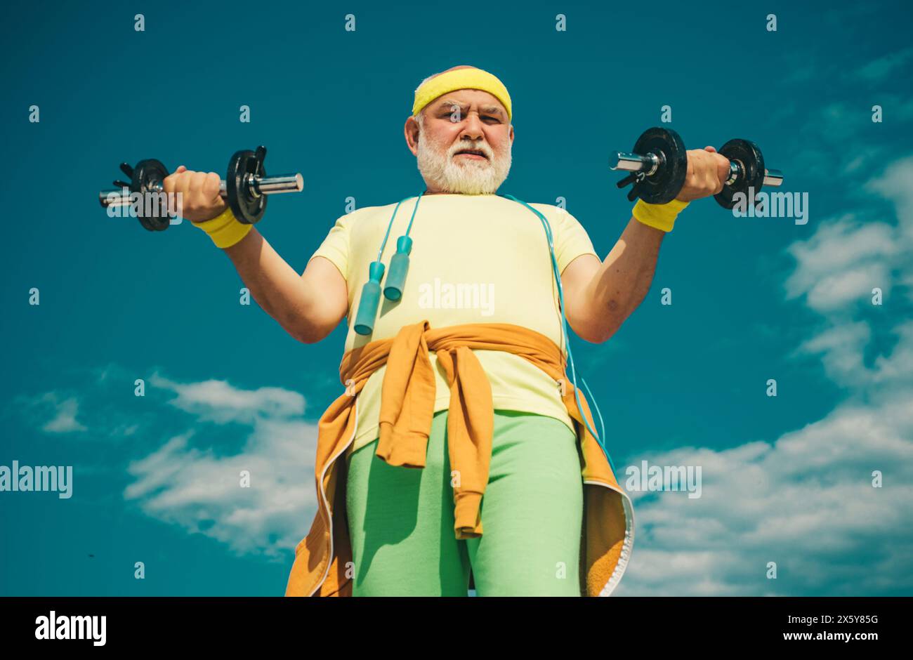 Senior sportman exercising with lifting dumbbell on blue sky background ...