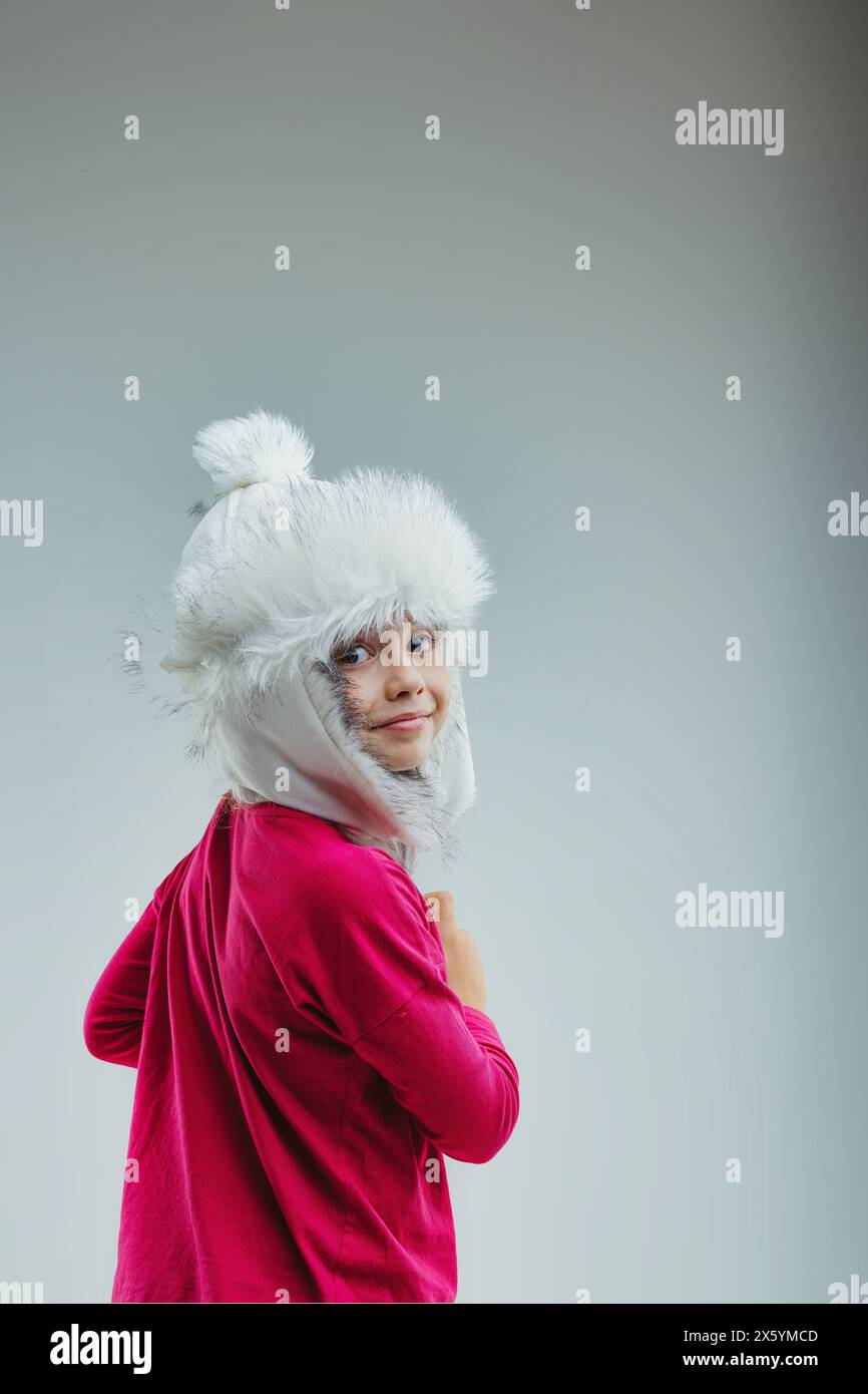 Young girl in a fluffy white winter hat and pink sweater glances over her shoulder with a ...