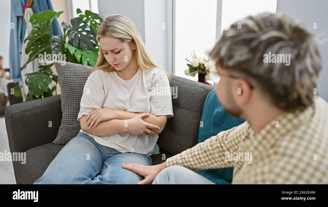 A distressed woman sits on a couch indoors as a concerned man reaches ...