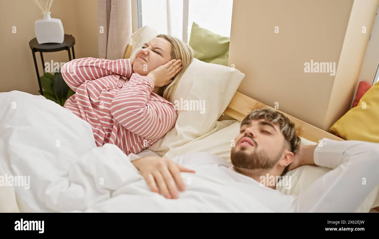 A young woman covers her ears beside a snoring man in a brightly lit ...