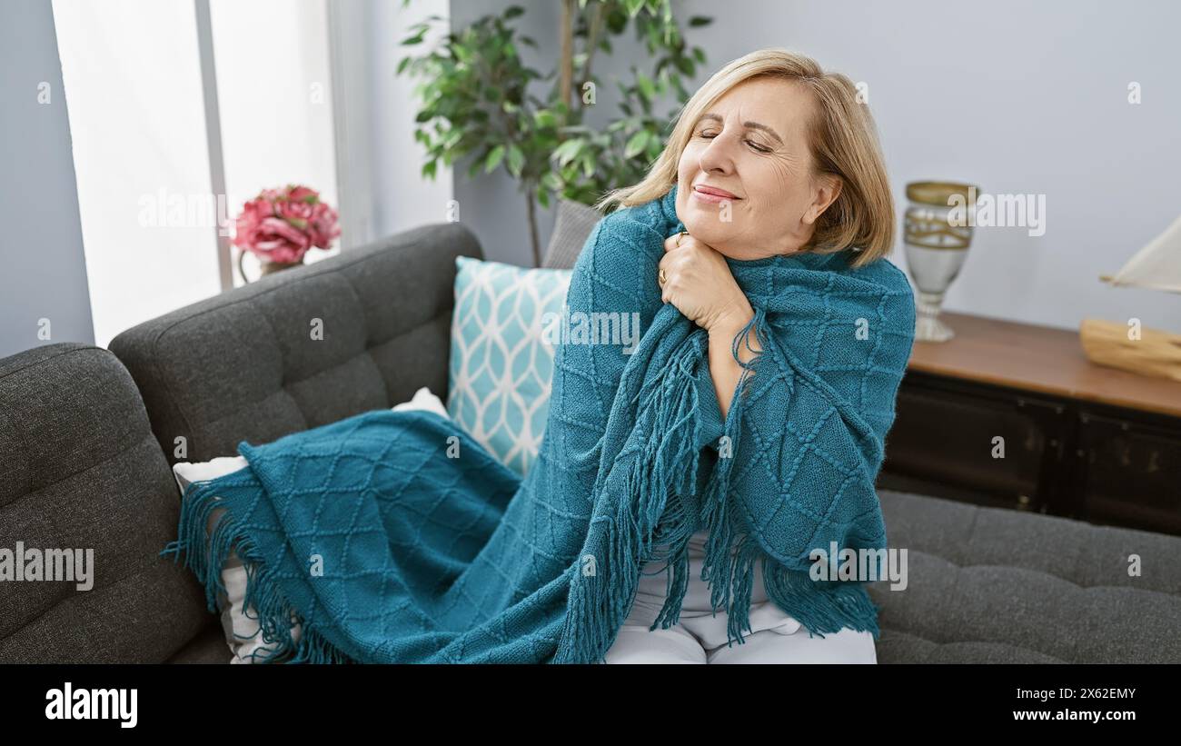 Blonde woman relaxing on a couch at home draped in a teal throw, eyes ...
