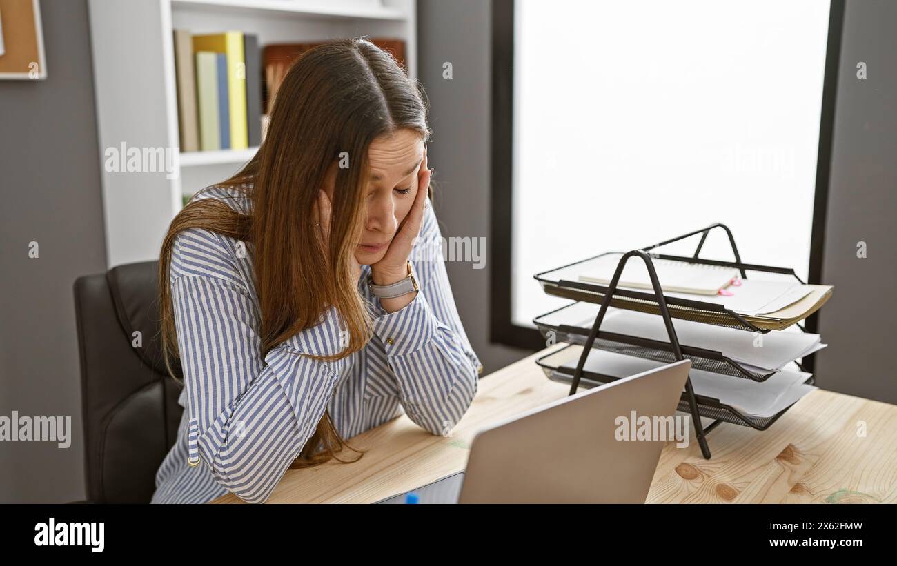 A stressed woman sits at her desk with a laptop in a modern office ...