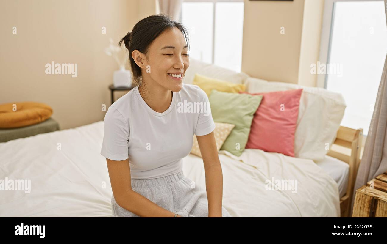 A cheerful asian woman sits on a bed in a well-lit, cozy bedroom ...