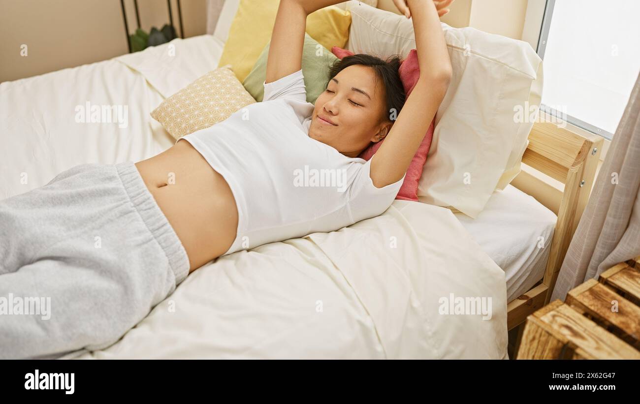 A relaxed asian woman stretching in a cozy bedroom setting, depicts ...