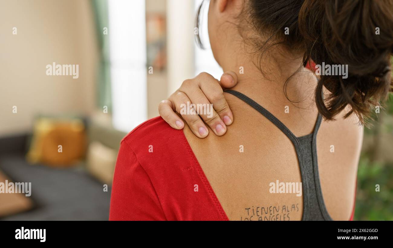 A young hispanic woman at home touches her shoulder, indicating back ...