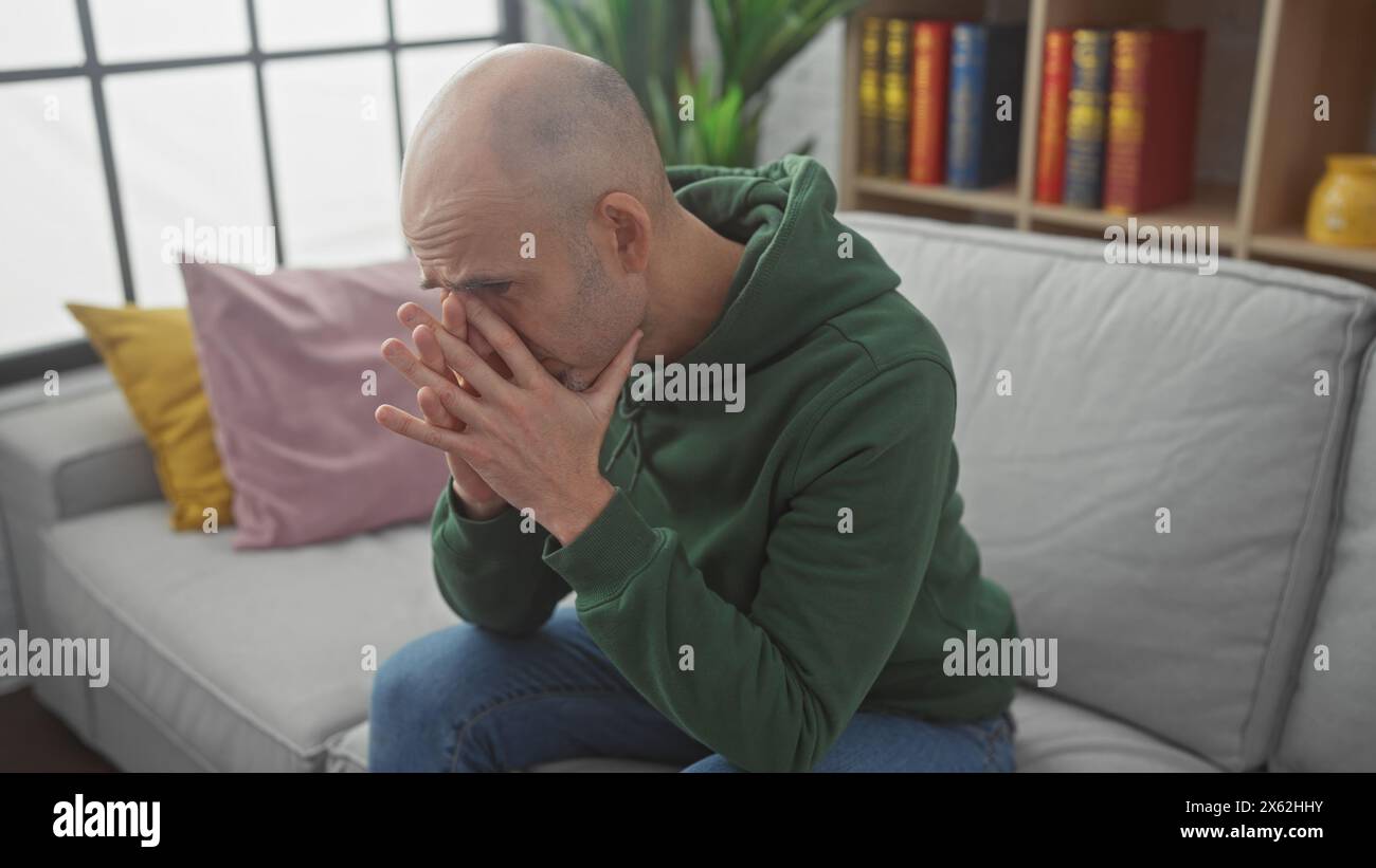A concerned bald man with a beard sitting on a couch indoors looking ...