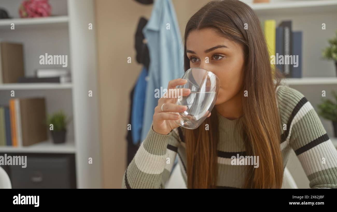 A young hispanic woman sips water in a modern indoor setting ...