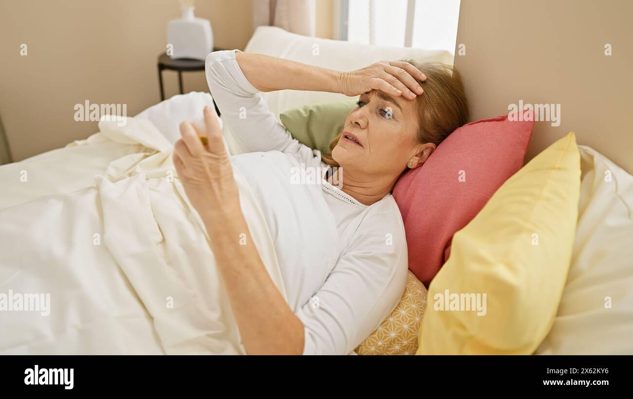 Mature blonde woman looks unwell holding medicine bottle in a bedroom ...