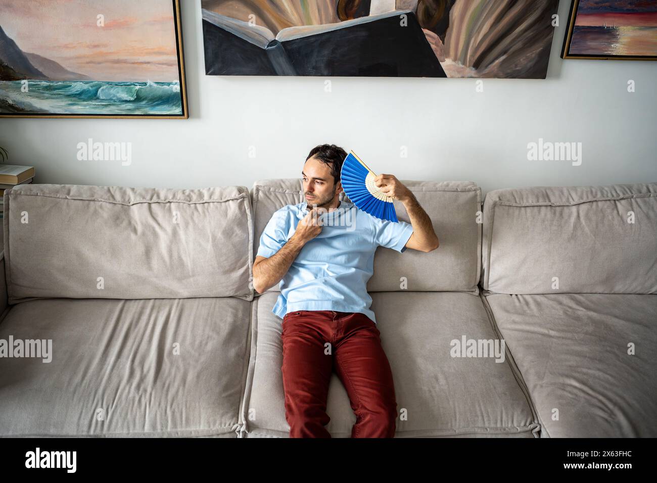 Guy coping with summer heat lying on sofa using paper fan suffocating ...