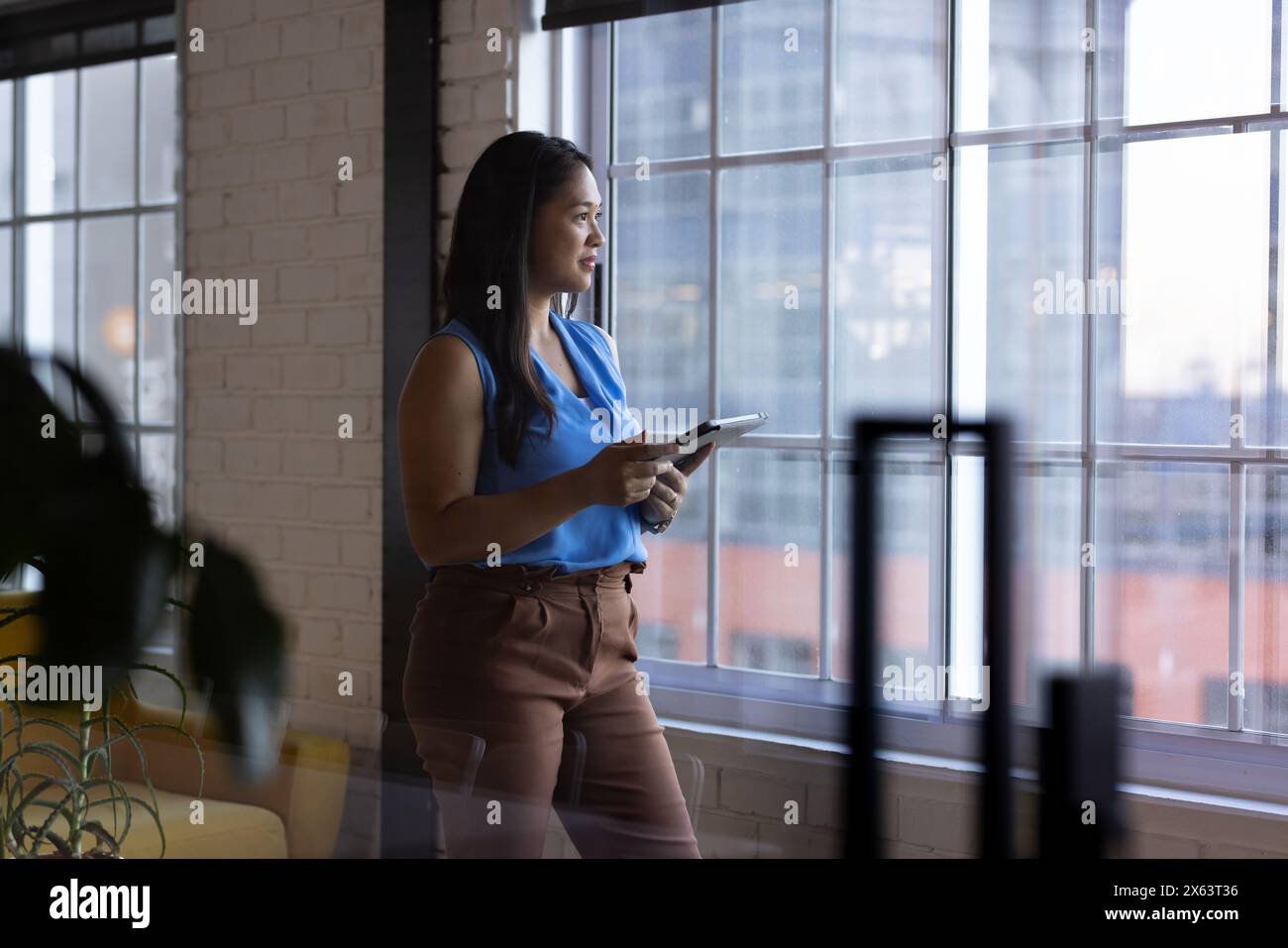 At office, biracial businesswoman holding tablet, gazing through window ...