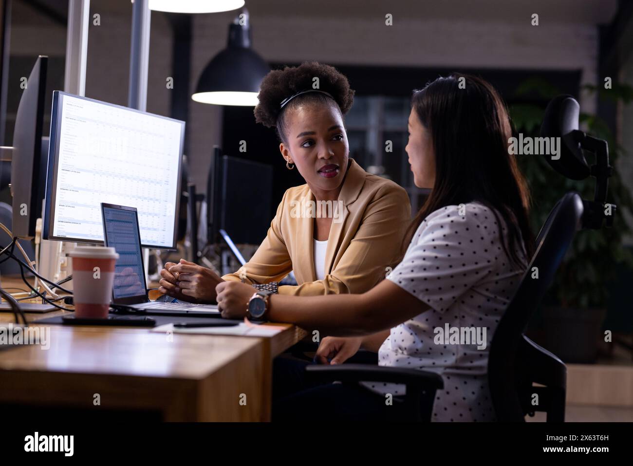 Diverse female colleagues discuss work late at office, using computers ...