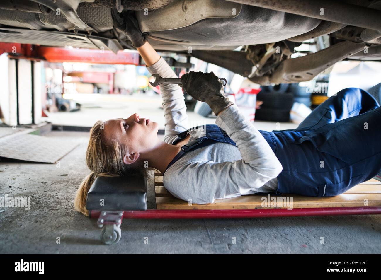 Female auto mechanic lying on mechanic creeper under car, inspecting and repairing vehicle ...