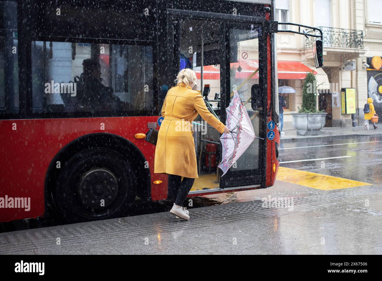 A woman closes her umbrella amidst the rain before boarding a city bus ...
