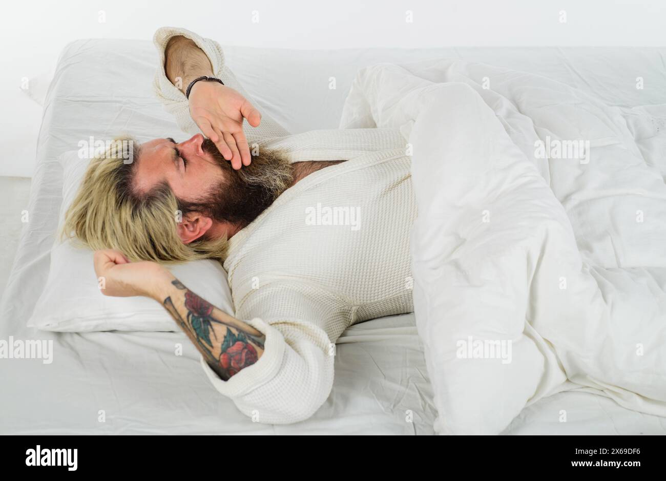 Bearded man in bathrobe lying on soft white pillow and yawning ...