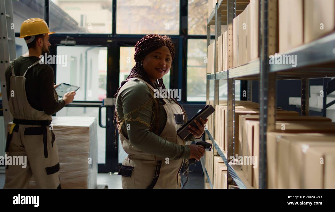 Dolly in portrait shot of cheerful african american warehouse employee scanning cardboard boxes ...