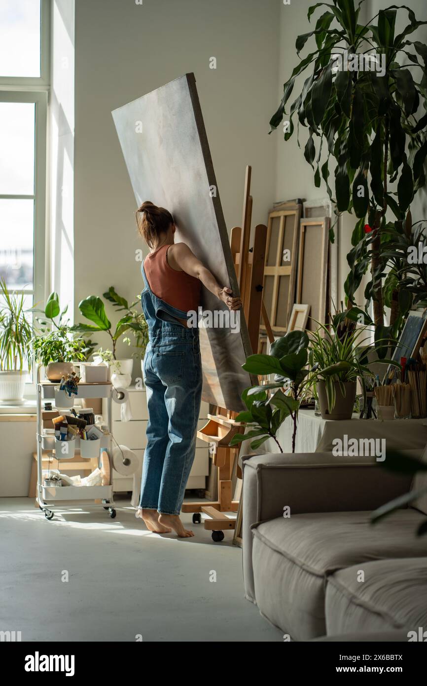 Interested artist young woman holds canvas puts on easel to continue ...