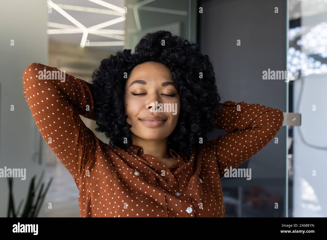 A young woman with closed eyes relaxing with hands behind her head in a modern office setting ...