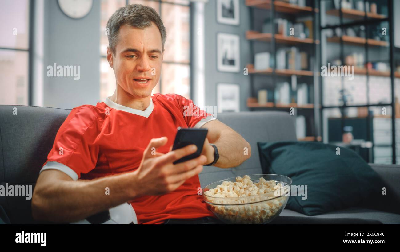 Charismatic Young Adult Man Sitting on a Couch Watches Game on TV, uses ...