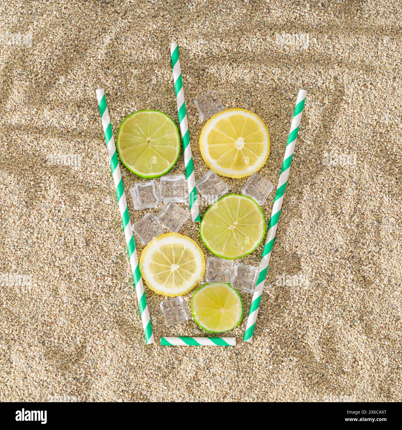 Creative summer composition with lemon and lime slices, drinking straw and ice cubes on sandy ...