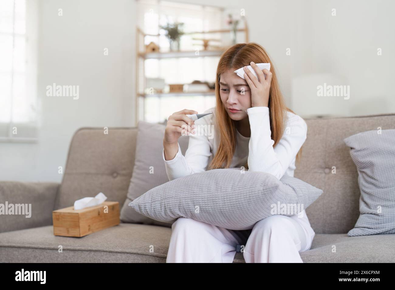 Sick young woman with fever checking thermometer and holding ice pack ...