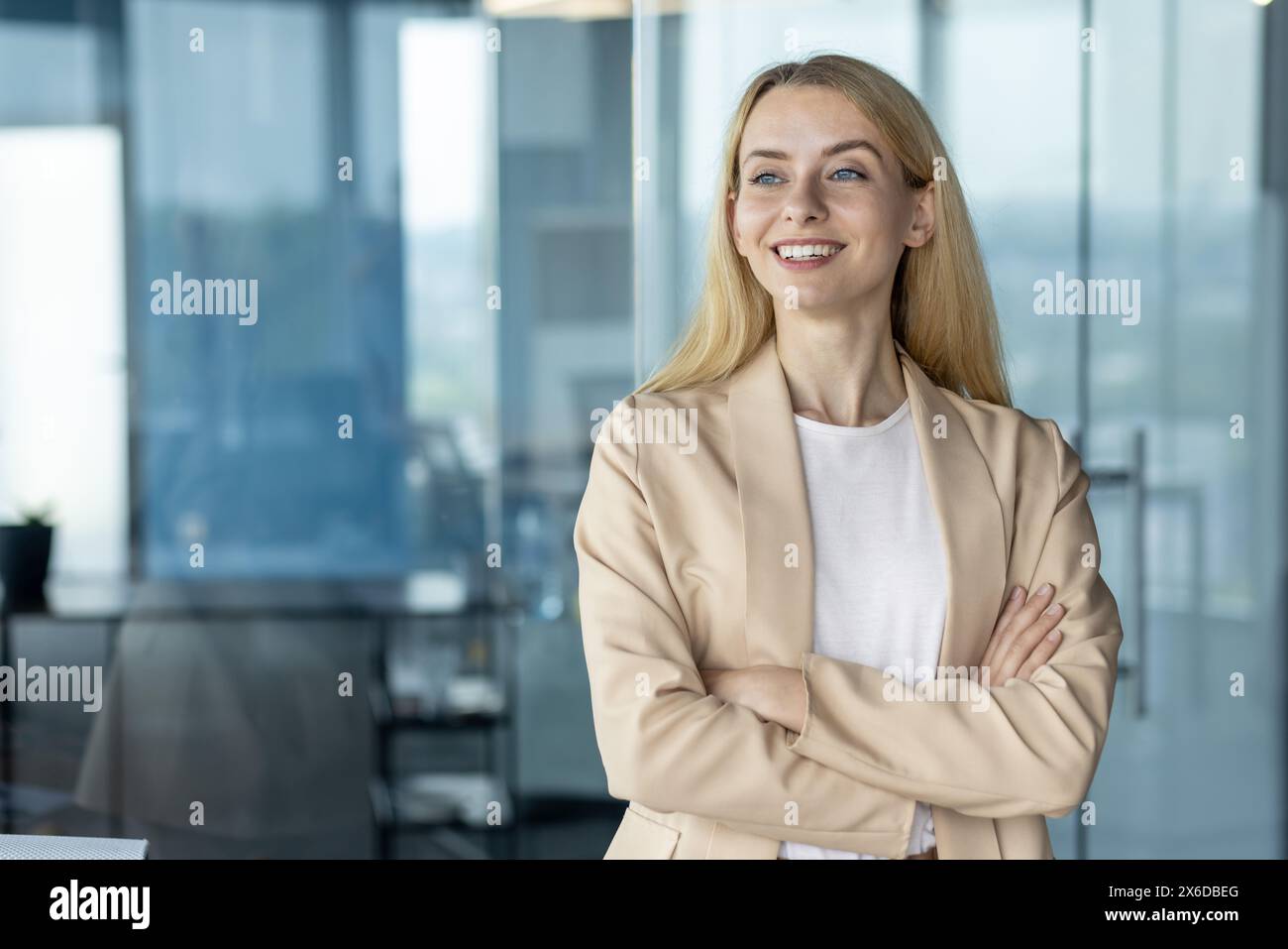 Portrait of a confident young businesswoman with arms crossed, smiling in a sunlit modern office ...