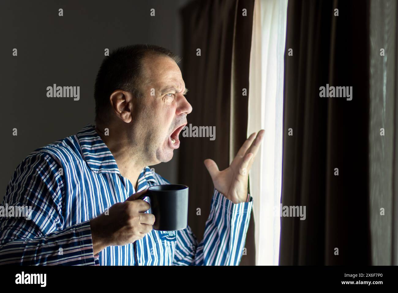 An angry man in striped pajamas with a mug of drink is looking out the ...
