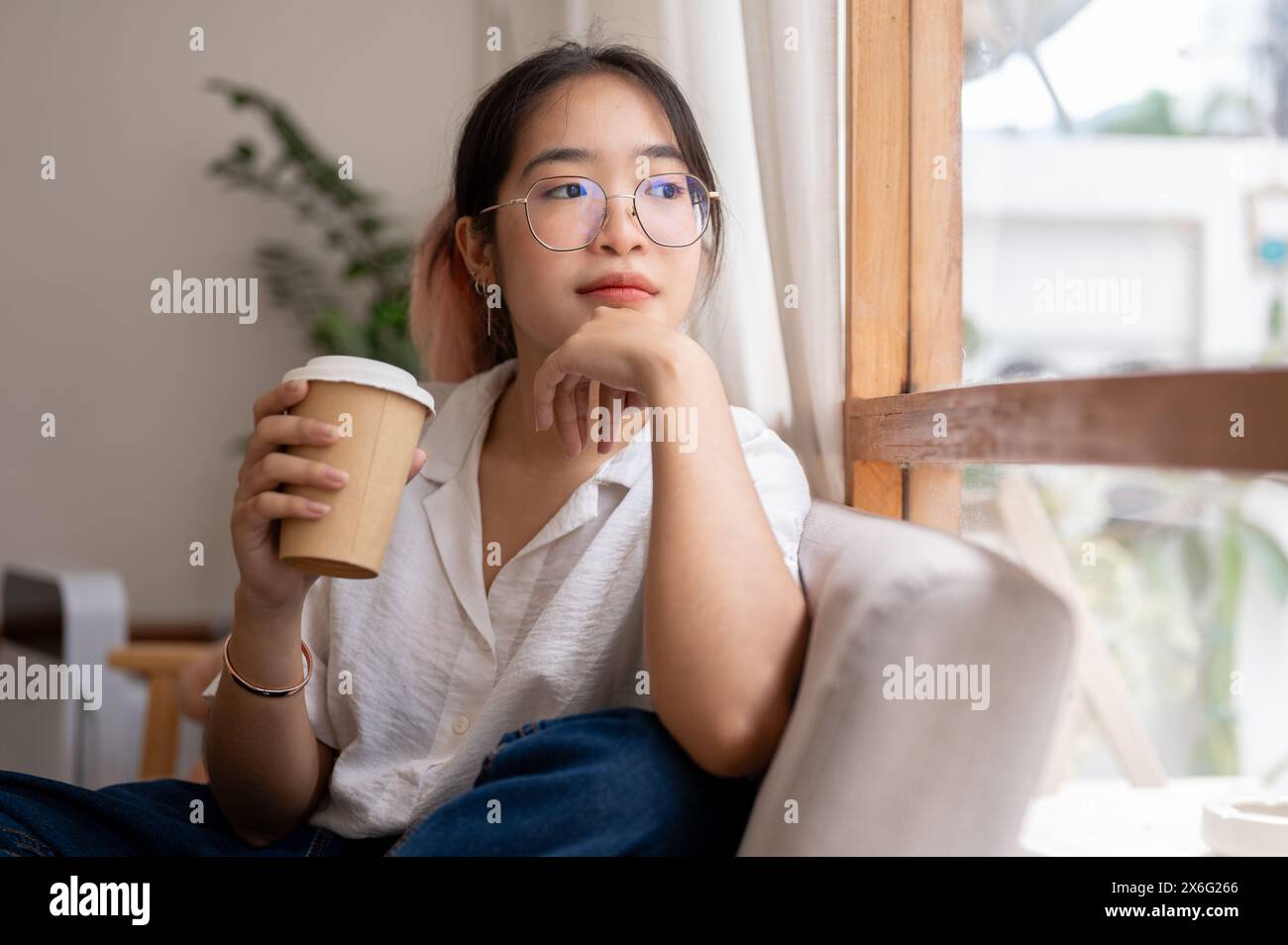 A young, thoughtful Asian woman is holding a coffee cup, looking out the window and pondering ...