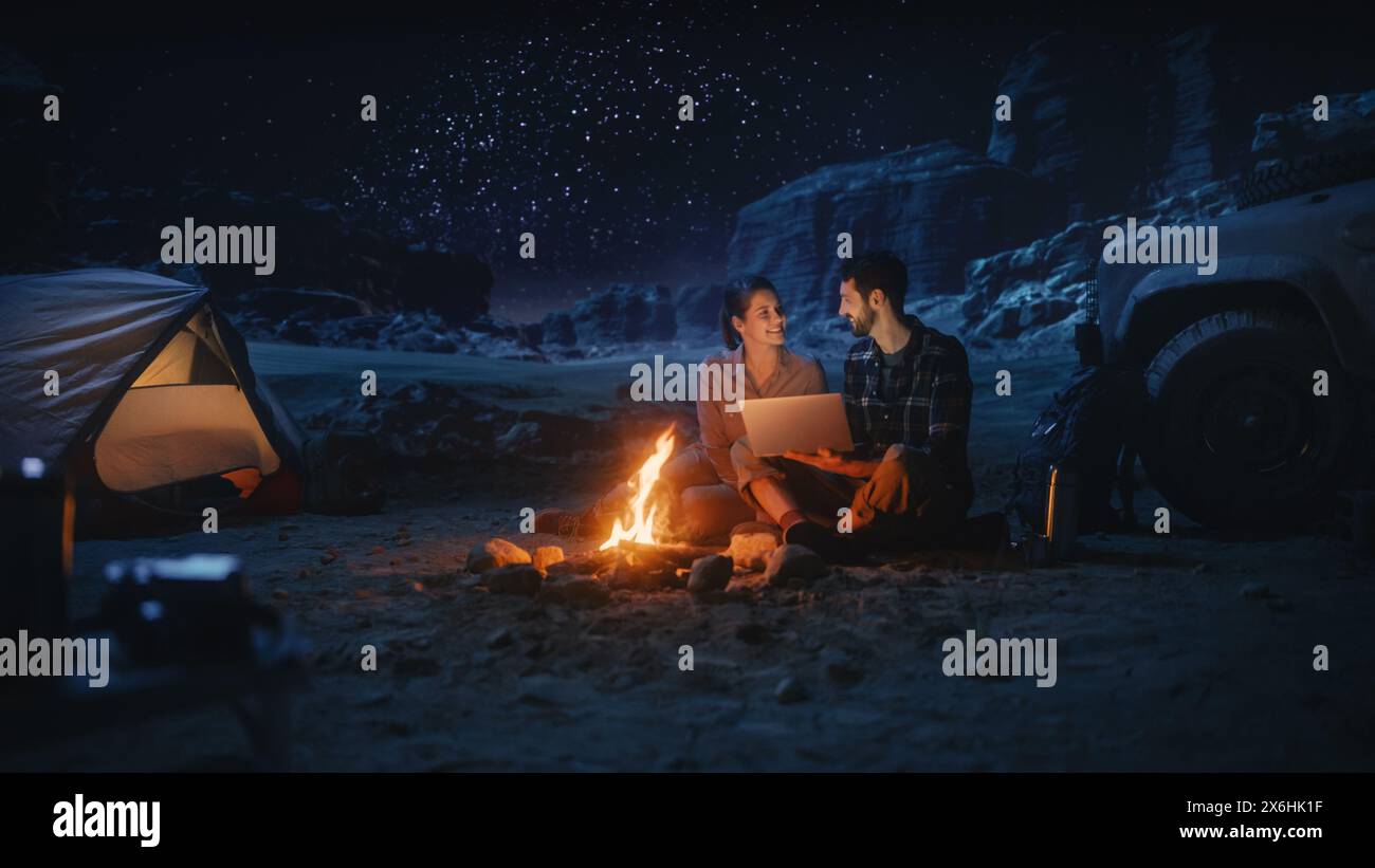 Happy Couple Camping in the Canyon at Night, Use Laptop Computer Sitting by Campfire. Two people ...
