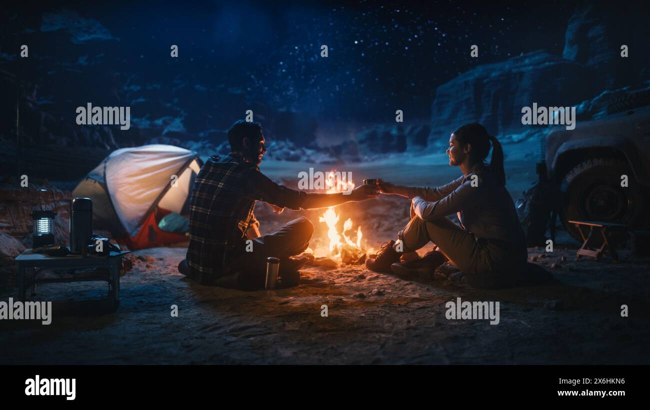 Happy Couple Sitting by Campfire Sharing Cup of Drink, Watching Night Sky while Camping in the ...