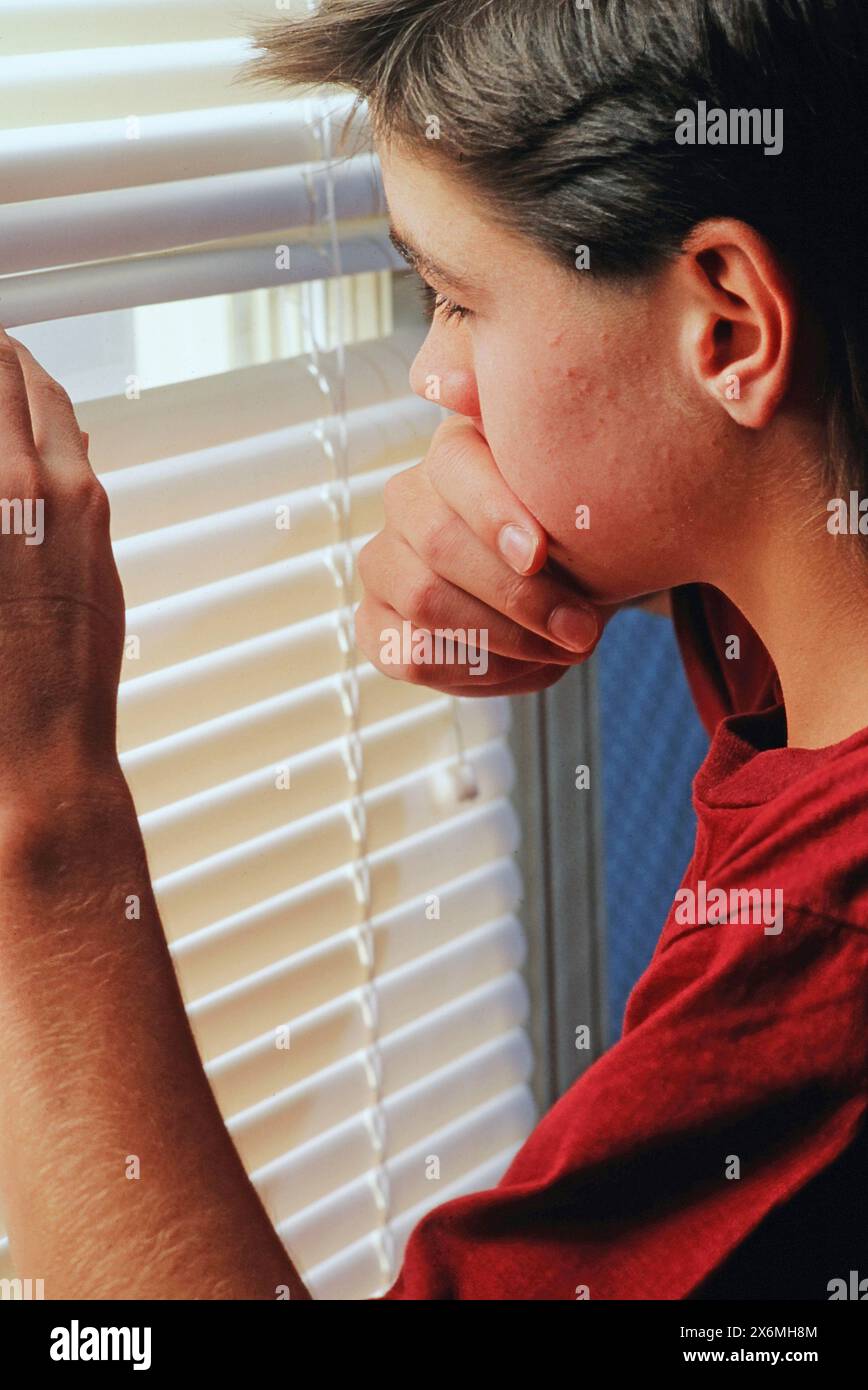 An anxious young teen boy is standing by a window with closed blinds ...